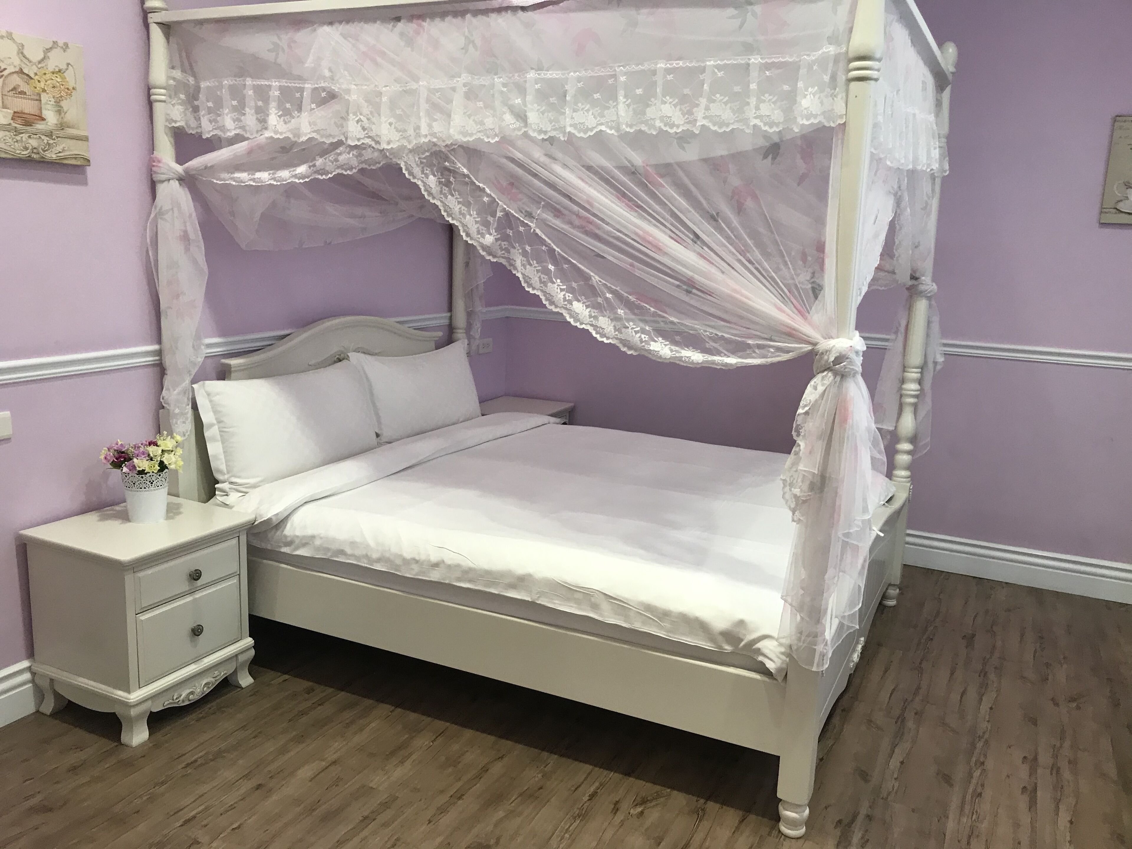 Uma cama com dossel e cortinas, acabamento branco, mesa de cabeceira com gavetas e um vaso com flores.