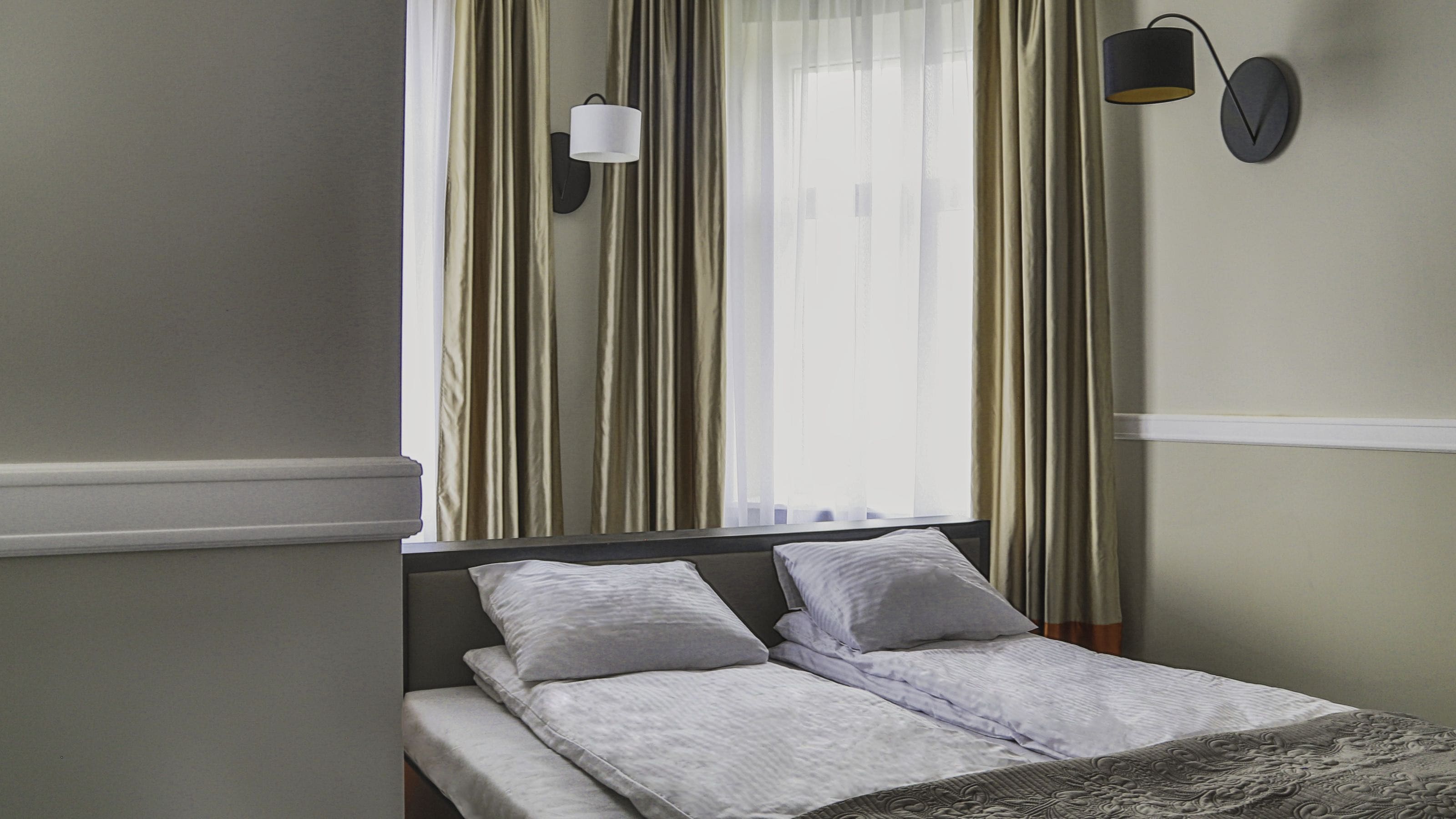 Uma cama de solteiro com duas almofadas, uma mesinha de cabeceira, uma luminária na parede e uma janela com cortinas.