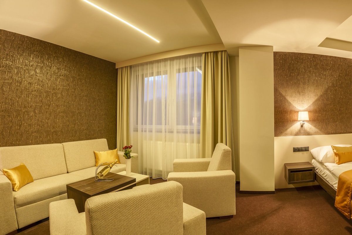 Quarto de hotel com sofá, poltronas, cama, mesa de cabeceira e luminária na parede.