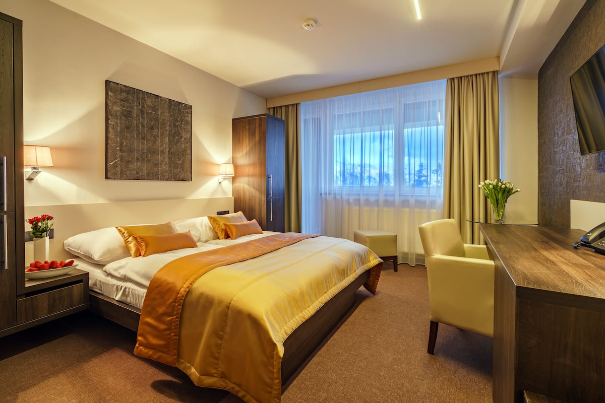 Quarto de hotel com uma cama grande, uma escrivaninha, uma cadeira, mesa de cabeceira com luminária e uma janela com cortinas.