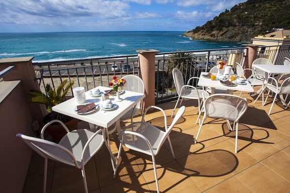 Terraço com cadeiras e mesas de metal branco, com vista para a praia e o oceano.