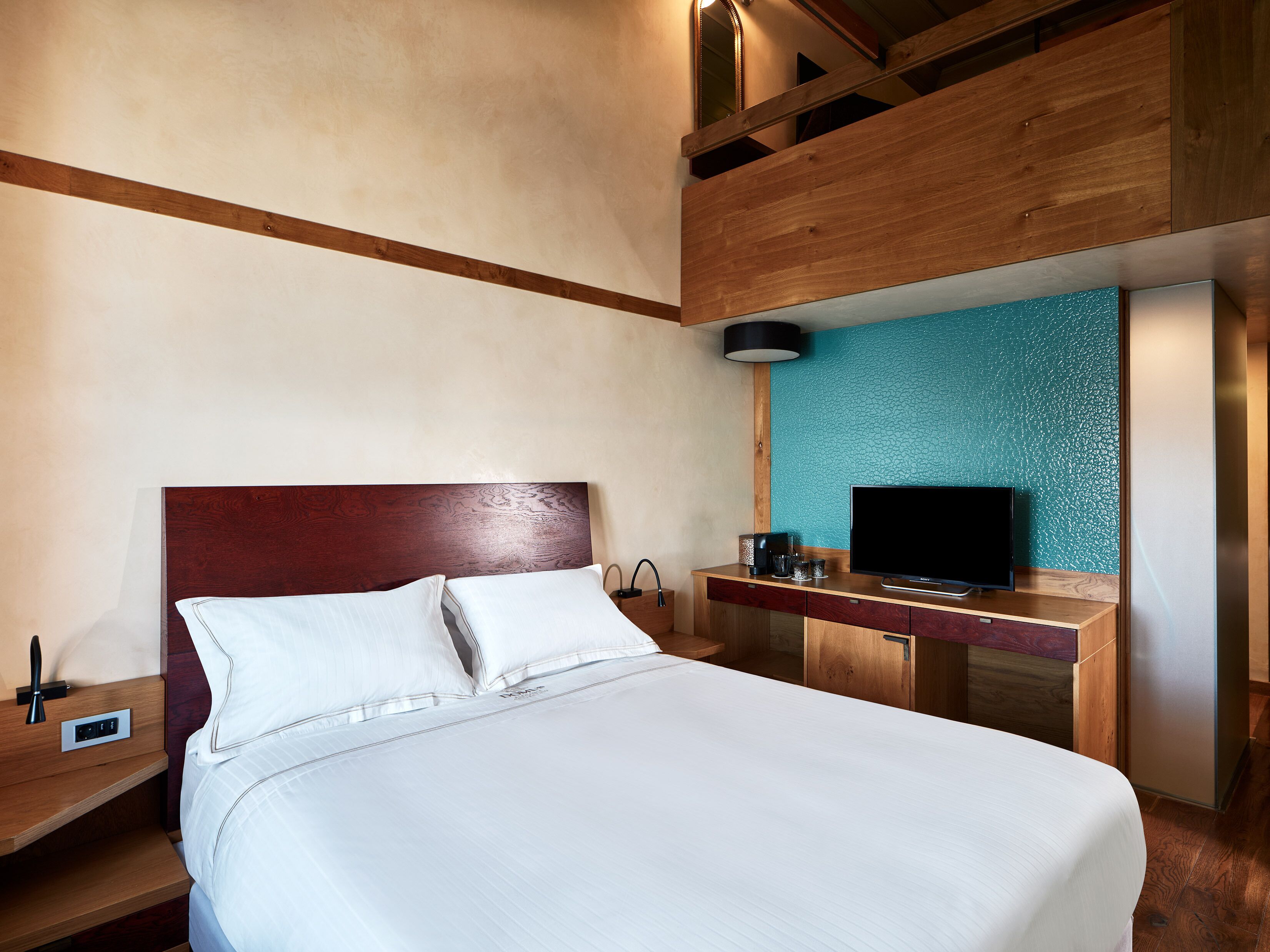 Quarto de hotel com uma cama grande, uma escrivaninha de madeira e uma televisão de tela plana, além de uma parede verde-água com textura.