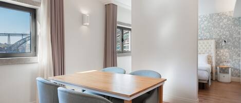 Quarto moderno com uma mesa de jantar de madeira, quatro cadeiras, uma cama com cabeceira branca, uma mesinha de cabeceira e vista para uma ponte através da janela.