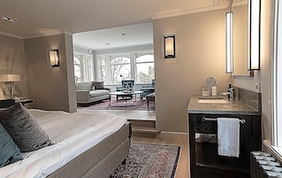 Quarto de hotel com cama, área de estar com sofá e cadeiras, banheiro com espelho e pia, e janelas amplas.