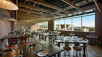 Um restaurante espaçoso com pé direito alto, vigas aparentes e grandes janelas que oferecem vista para a paisagem urbana.