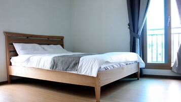 Uma cama de solteiro com estrutura de madeira e roupa de cama branca num quarto com chão de madeira claro e uma janela com cortinas azuis.