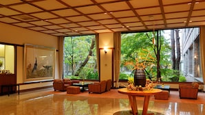 Um hall amplo com uma grande janela que oferece vista para árvores e um edifício. Há vários espaços de estar com sofás e uma mesa central com um vaso.