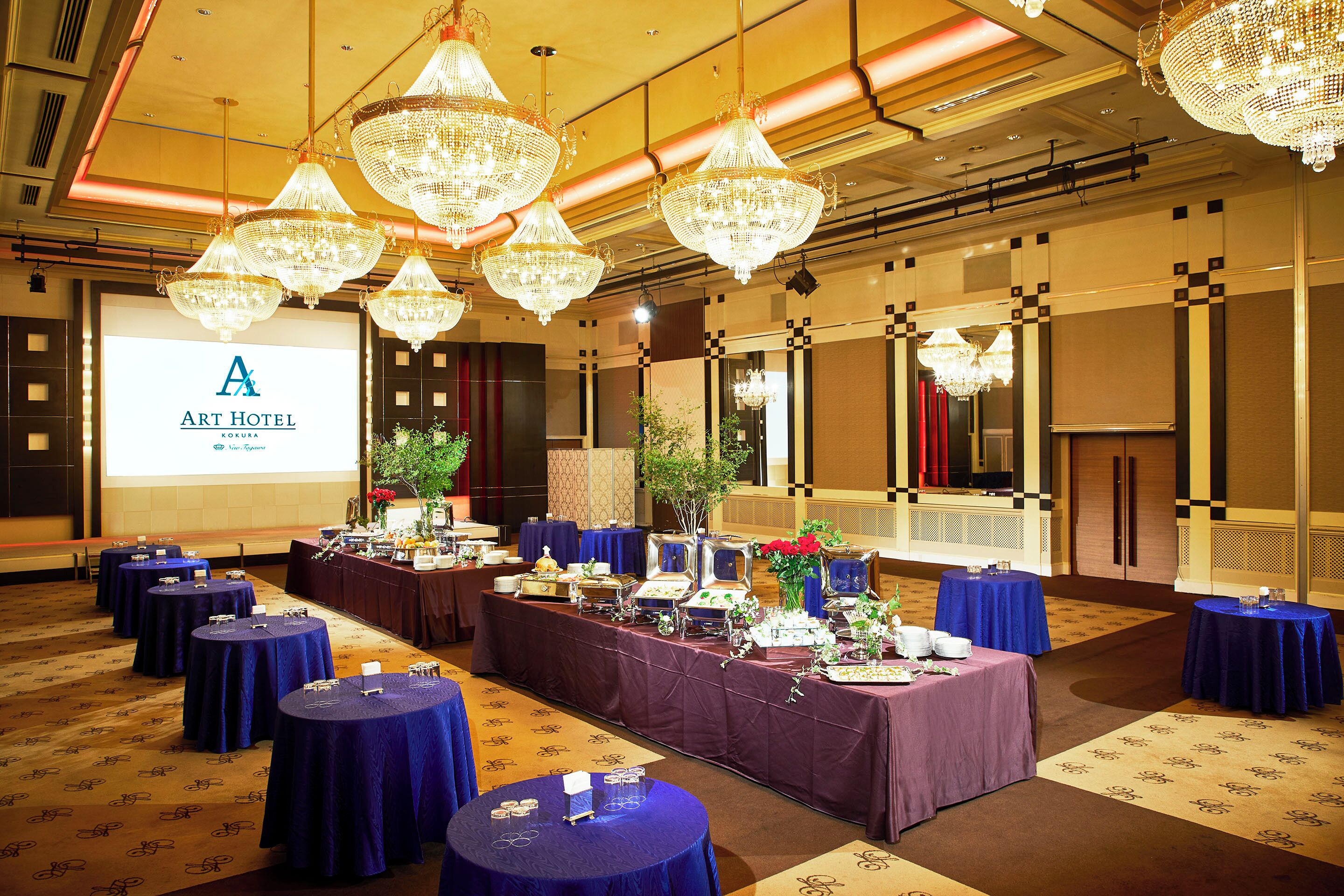 Uma sala de banquetes com várias mesas redondas, uma tela grande exibindo o logotipo do Art Hotel e luminárias de cristal elaboradas.