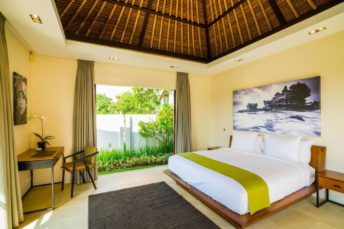 Quarto com uma cama grande, uma escrivaninha e uma cadeira. Há uma janela com cortinas, um quadro na parede e vista para o verde lá fora.