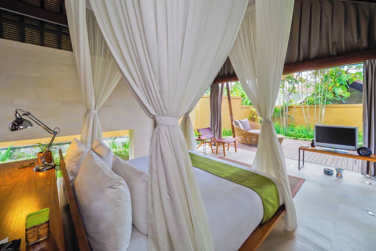 Quarto com cama de dossel, mesa de cabeceira com abajur e vista para o pátio externo.