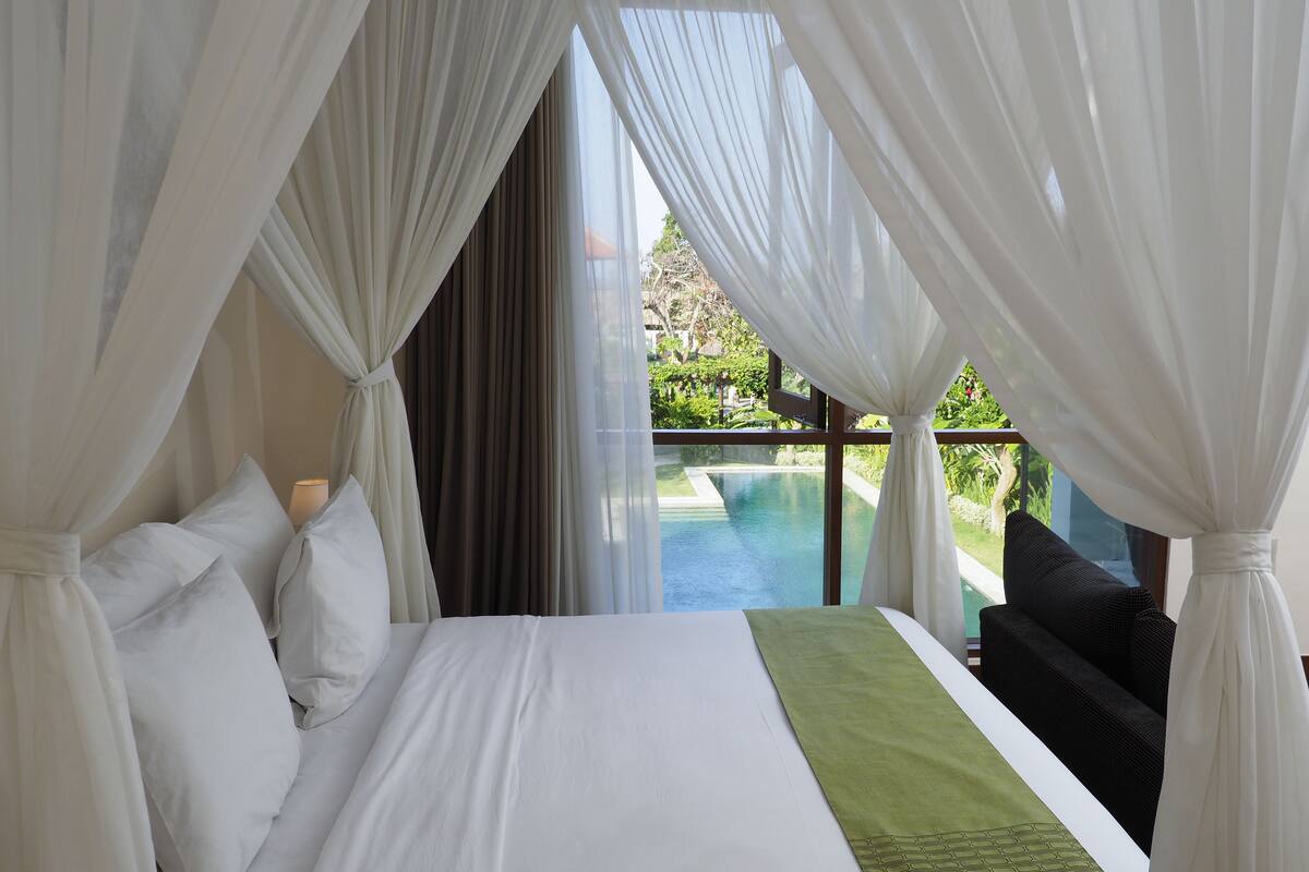Quarto com cama grande, cortinas brancas e vista para uma piscina ao ar livre.