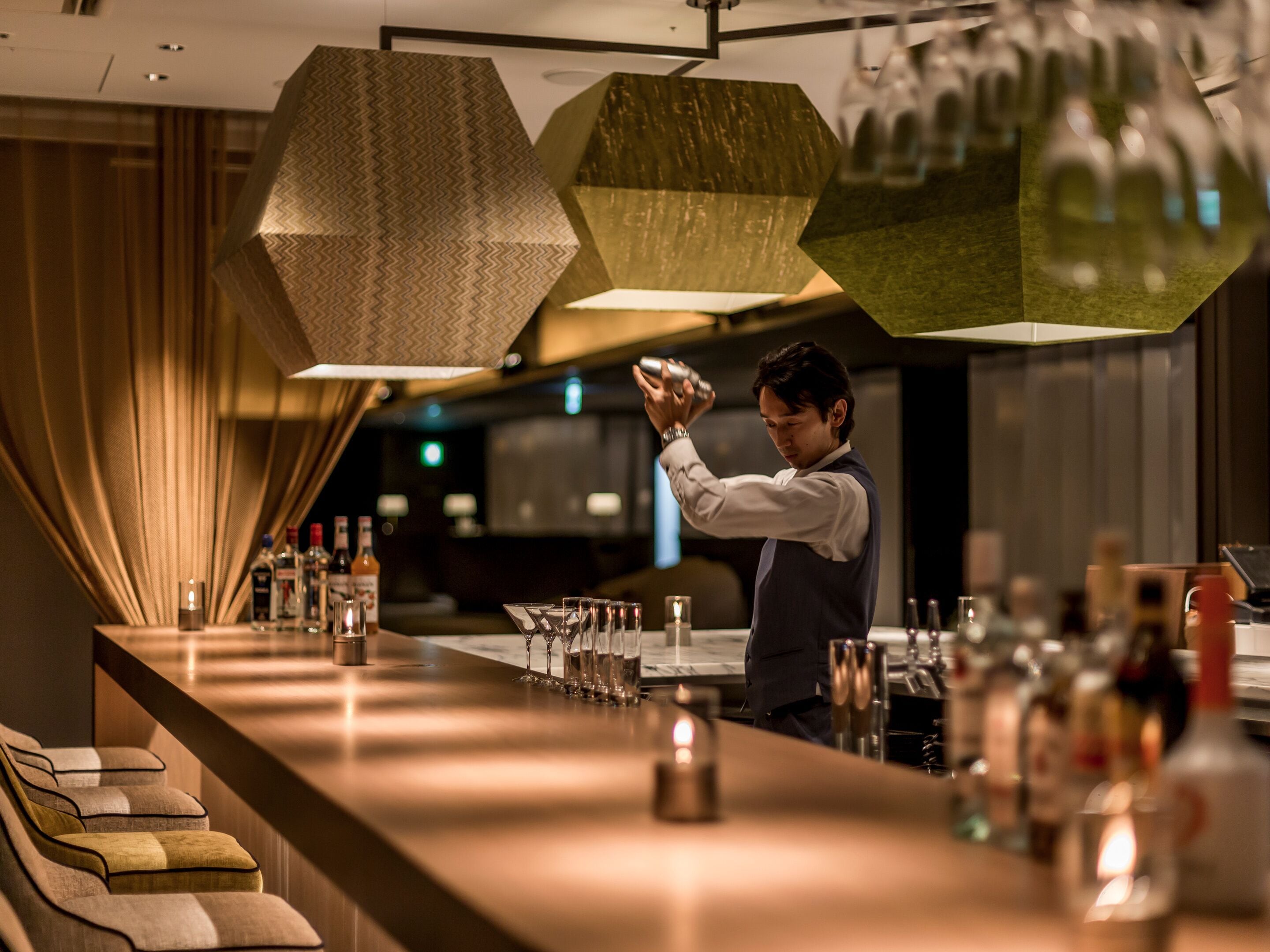 Uma área de bar com um bartender preparando uma bebida, banquetas e um balcão longo com garrafas e copos.
