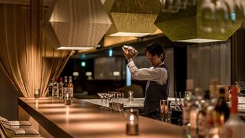 Uma área de bar com um bartender preparando uma bebida, banquetas e um balcão longo com garrafas e copos.