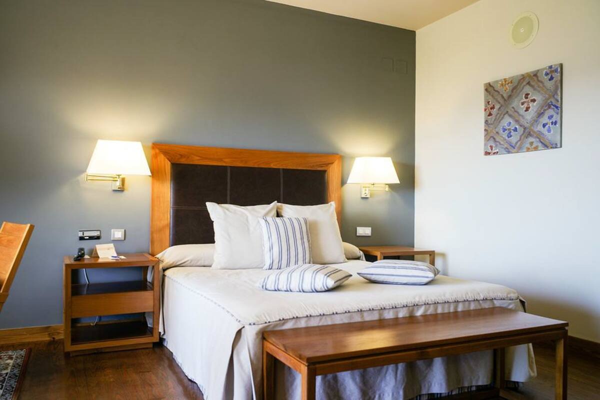 Quarto bem arrumado com uma cama grande, duas mesas de cabeceira, uma mesinha de madeira, um banquinho e uma luminária na parede.