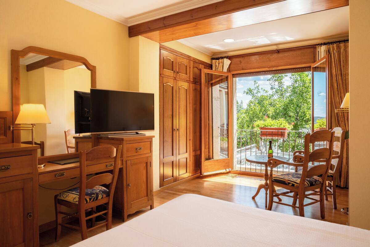 Quarto de hotel com cama, mobiliário de madeira, televisão e varanda com mesa e cadeiras.