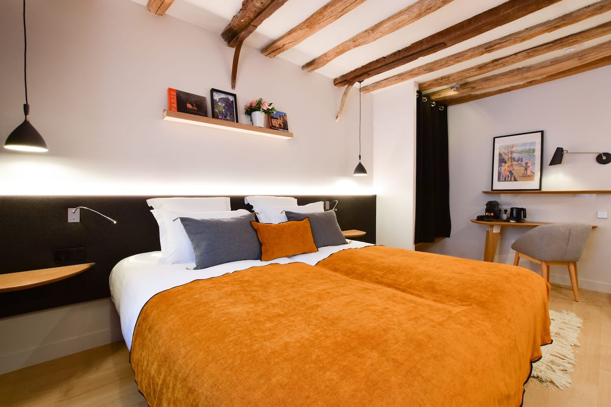 Quarto com uma cama grande, vigas de madeira, prateleira com fotos e uma planta, escrivaninha com cadeira e quadro pendurado na parede.