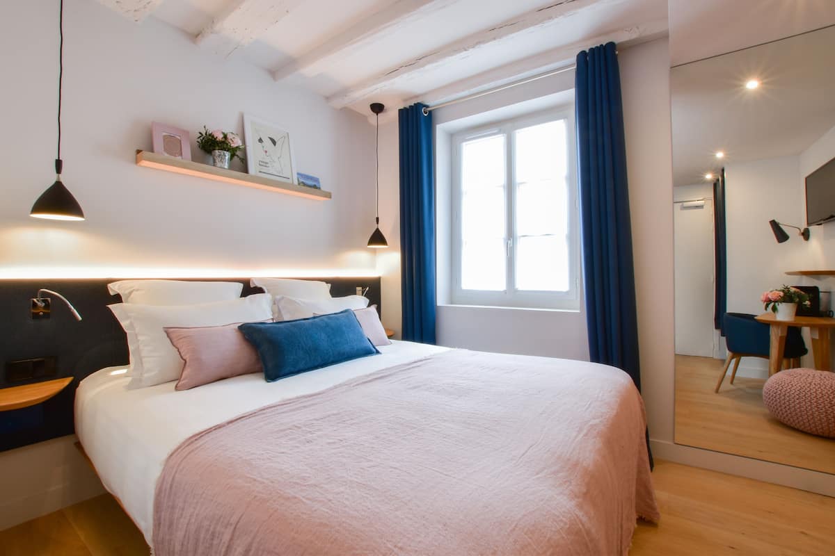 Quarto arrumado com uma cama grande, mesas de cabeceira, luminária, janela com cortinas, espelho e estante com objetos decorativos.