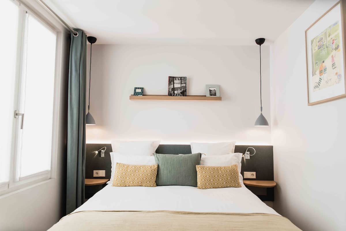 Um quarto bem arrumado com uma cama, mesas de cabeceira, uma estante com livros e fotografias, e uma janela com cortinas.