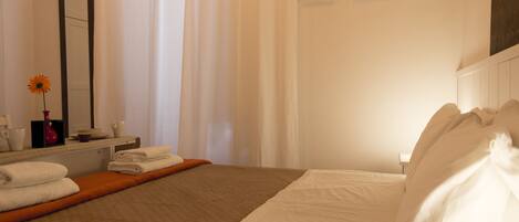 Quarto de hotel com cama, espelho, toucador com artigos de higiene e janela com cortinas.