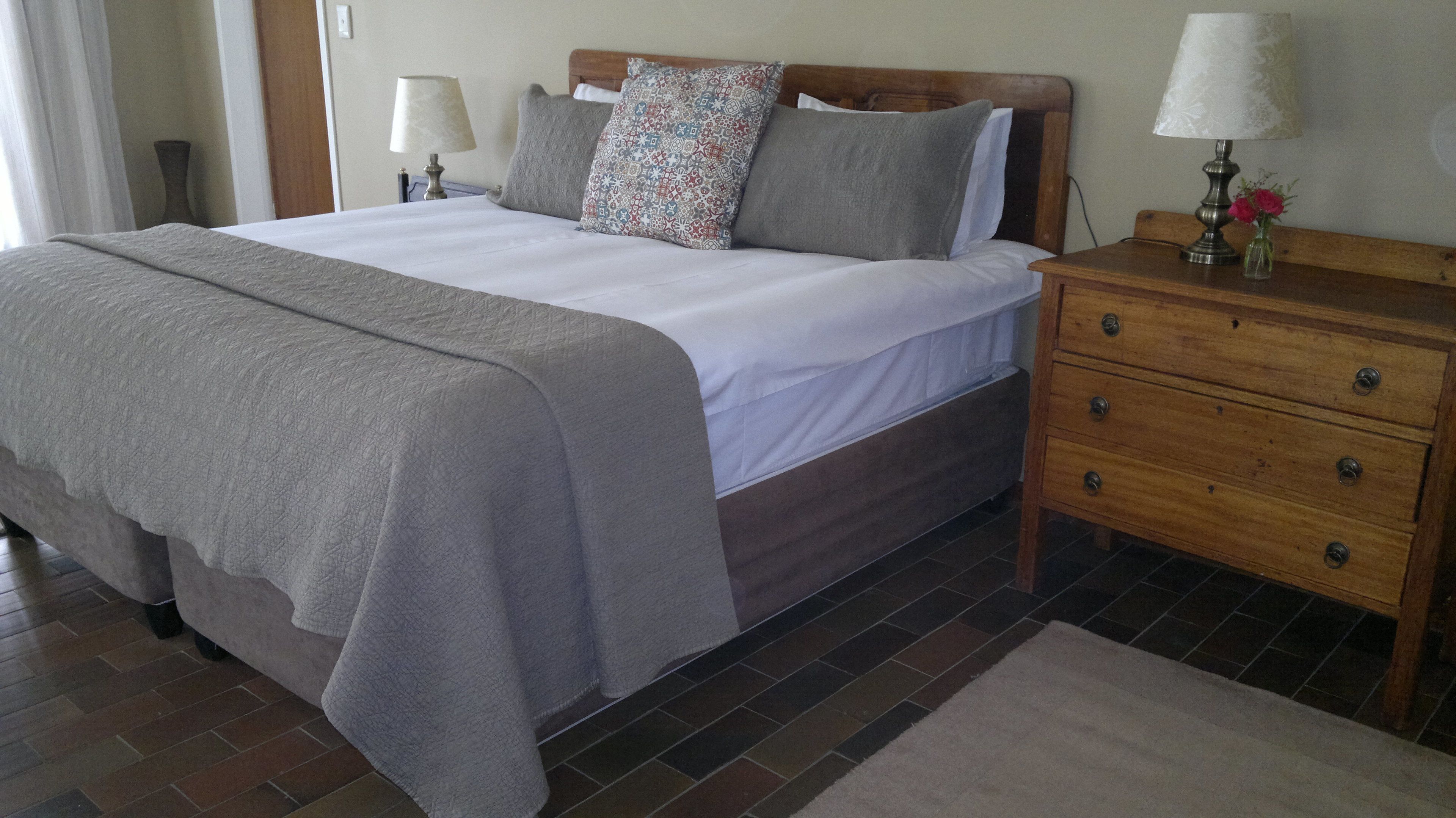 Uma cama bem arrumada com um cobertor cinza, uma mesa de cabeceira de madeira com gavetas e um vaso com flores.