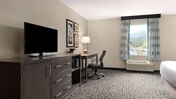Quarto de hotel com televisão de tela plana, uma escrivaninha com cadeira, luminária, cama e janela com cortinas.