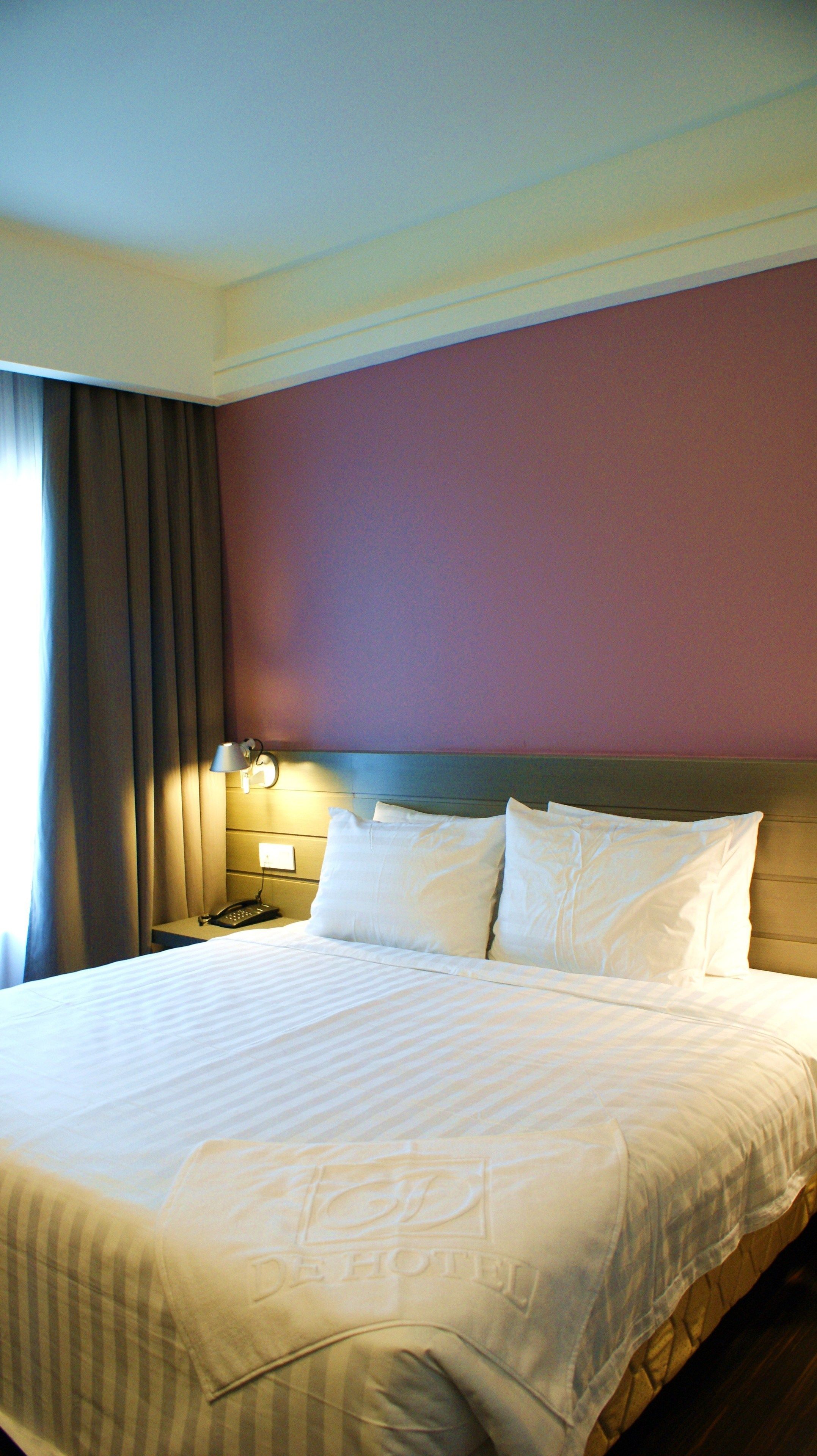 Quarto de hotel com uma cama grande, uma mesa de cabeceira, uma luminária e uma janela com cortinas.