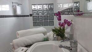 Casa de banho com uma pia branca, um espelho e uma parede decorada com espelhos quadrados. Existem duas toalhas brancas no chão e um vaso com flores roxas na pia.