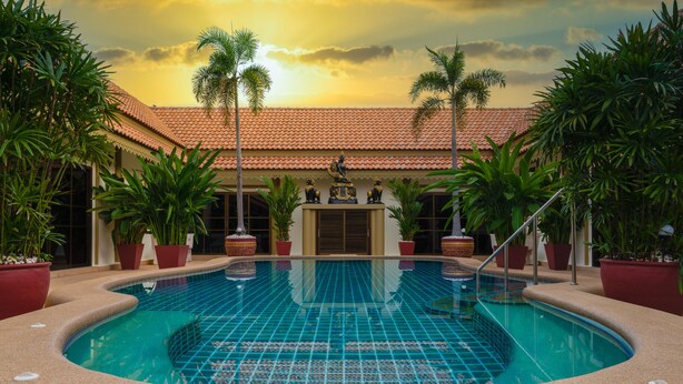 Piscina com chão em azulejos, cercada por plantas em vasos e palmeiras, com um edifício com telhado em azulejos ao fundo.