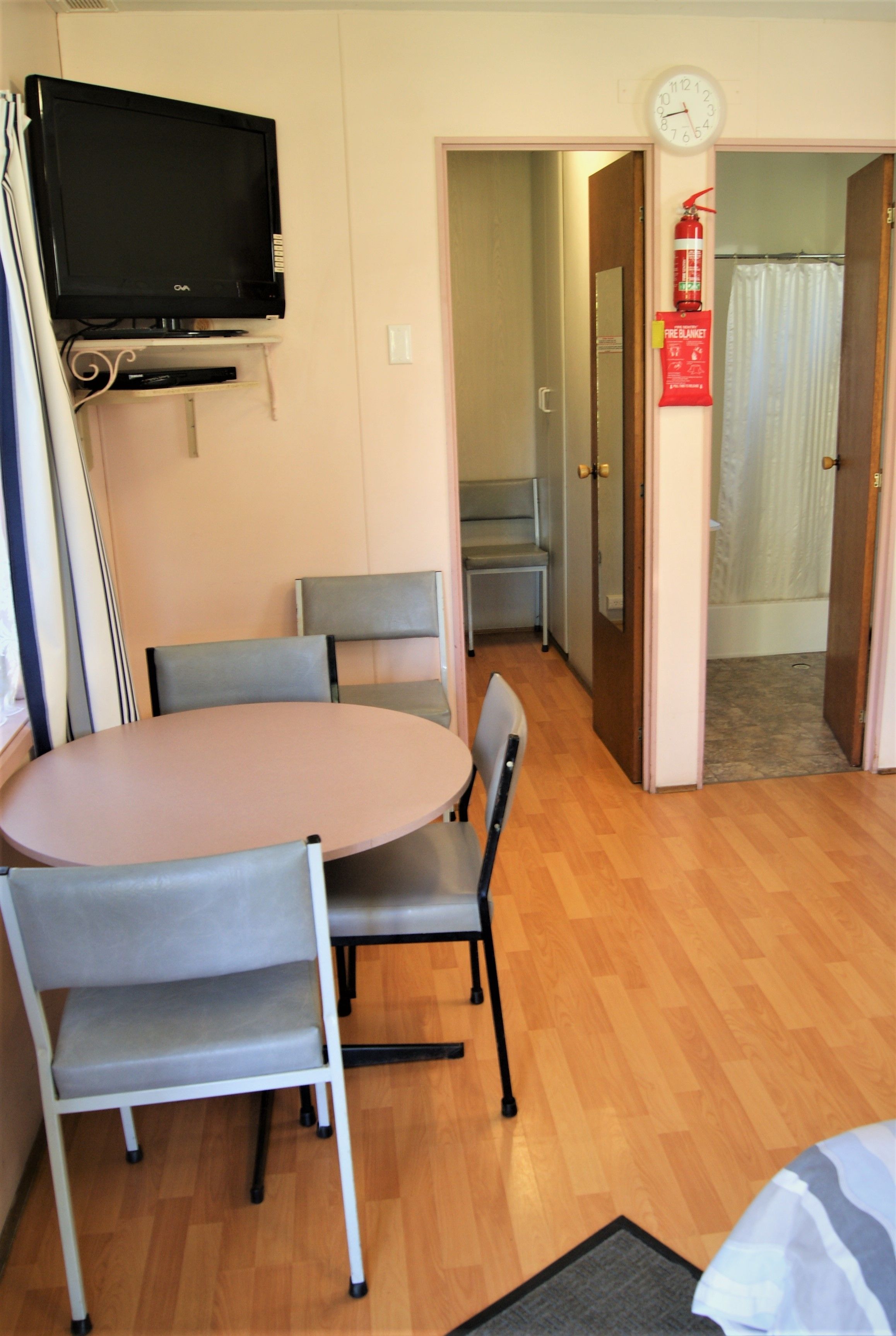 Um quarto com uma mesa redonda, quatro cadeiras, uma televisão fixada na parede, um extintor de incêndio e um relógio.
