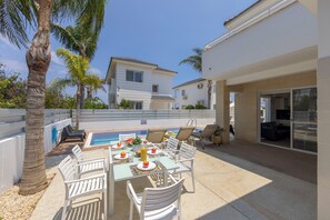 Uma área de refeições à beira da piscina, com uma mesa posta para dois, cercada por cadeiras, piscina e palmeiras.