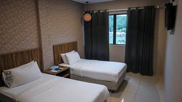 Quarto de hotel com duas camas, cabeceira de madeira, mesa de cabeceira, janela com cortinas e vista para o exterior.