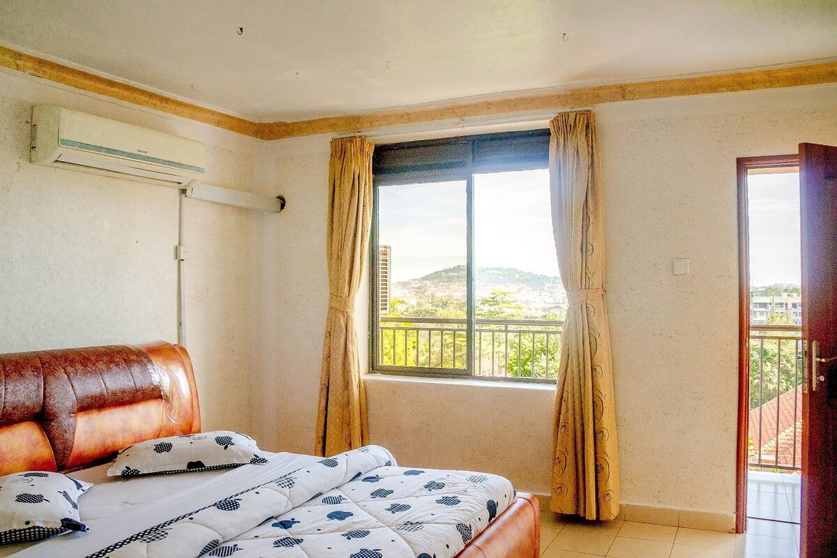 Quarto com cama, janela com cortinas, vista para vegetação e porta para varanda.