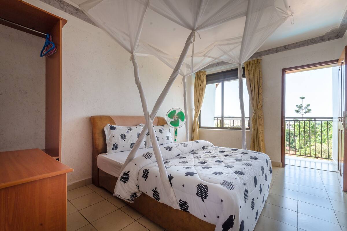 Quarto com cama, mesa de cabeceira, ventoinha e varanda com vista.