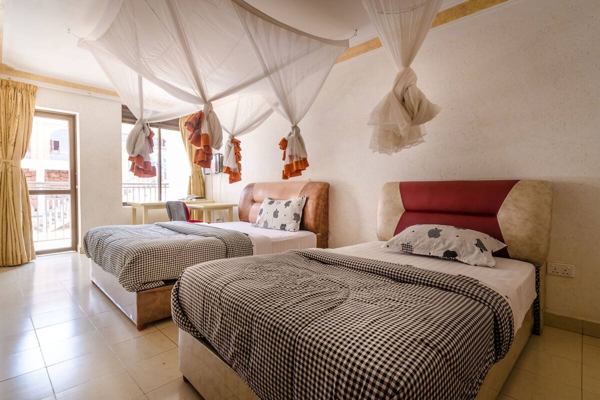 Quarto de hotel com duas camas, uma escrivaninha, uma cadeira e uma janela com cortinas.