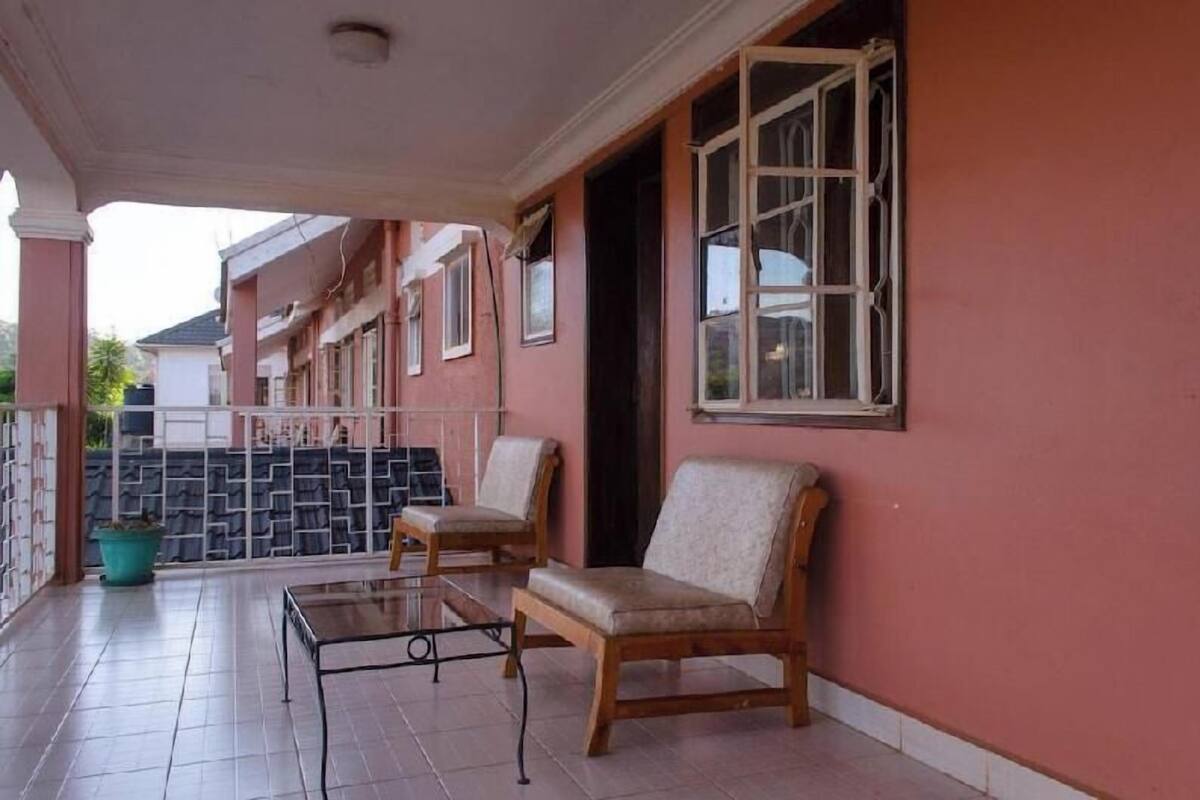 Uma área externa coberta com duas cadeiras de madeira, uma mesa de vidro, piso de azulejos e uma parede rosa com janelas.