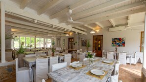 Uma sala de jantar ampla com mesas e cadeiras cobertas de branco, janelas grandes e teto rústico.