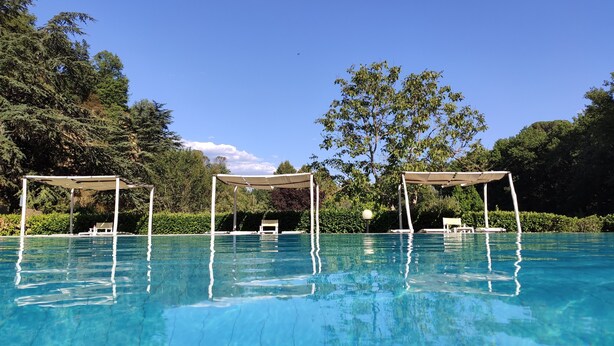 Piscina cristalina com três áreas de estar à sombra e vegetação exuberante ao fundo.
