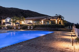 Piscina com espreguiçadeiras e guarda-sóis, área de estar coberta ao ar livre e um cenário de encosta.