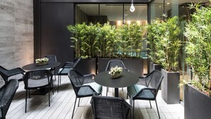 Uma área moderna para refeições ao ar livre, com cadeiras de vime preto e uma mesa redonda, cercada por vegetação exuberante e uma parede escura.