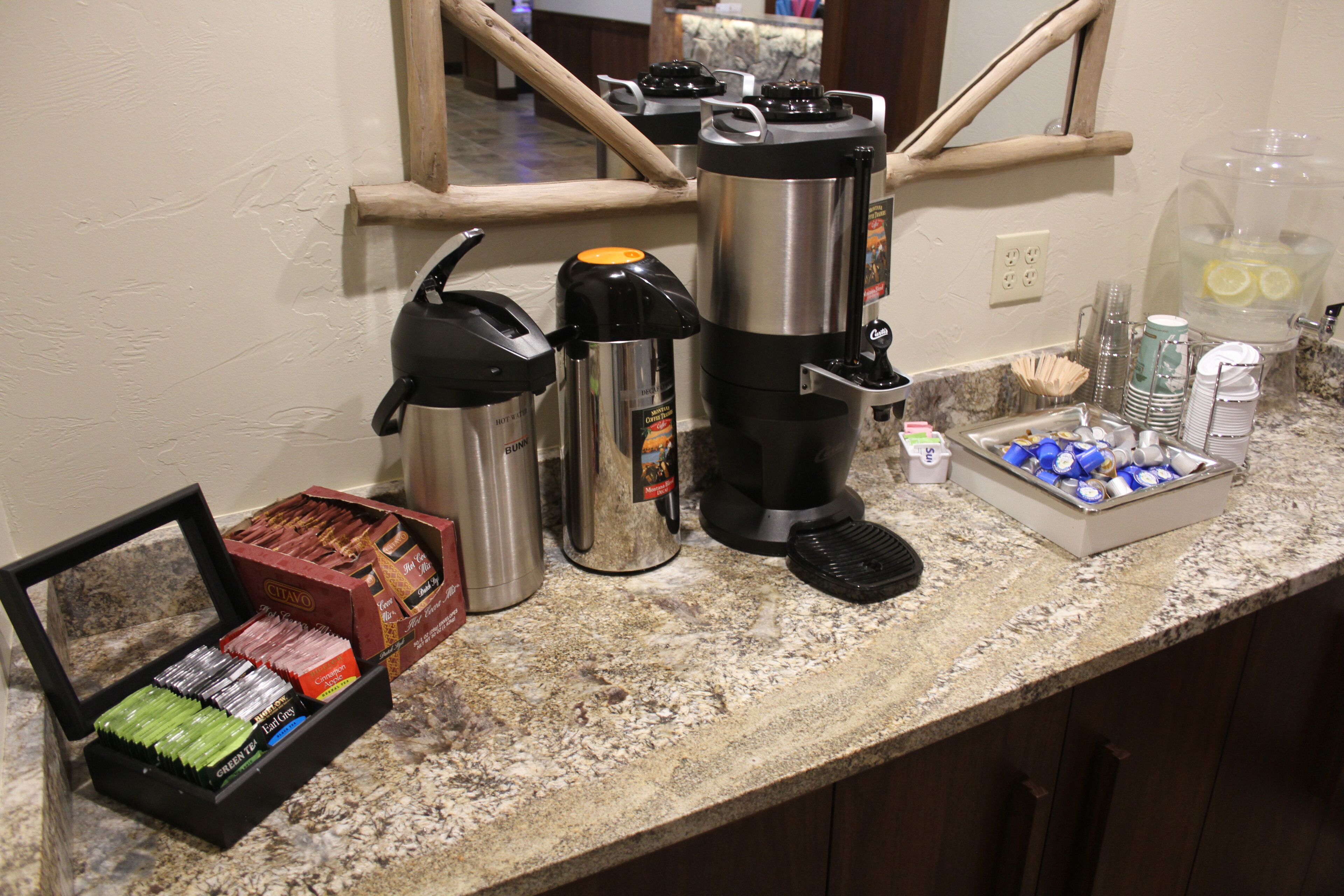 Uma bancada de cozinha com cafeteira, um termo, um bebedouro e uma caixa de saquinhos de chá.