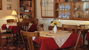 Uma área de refeições com mesas e cadeiras de madeira, um vaso com flores e uma estante de livros ao fundo.