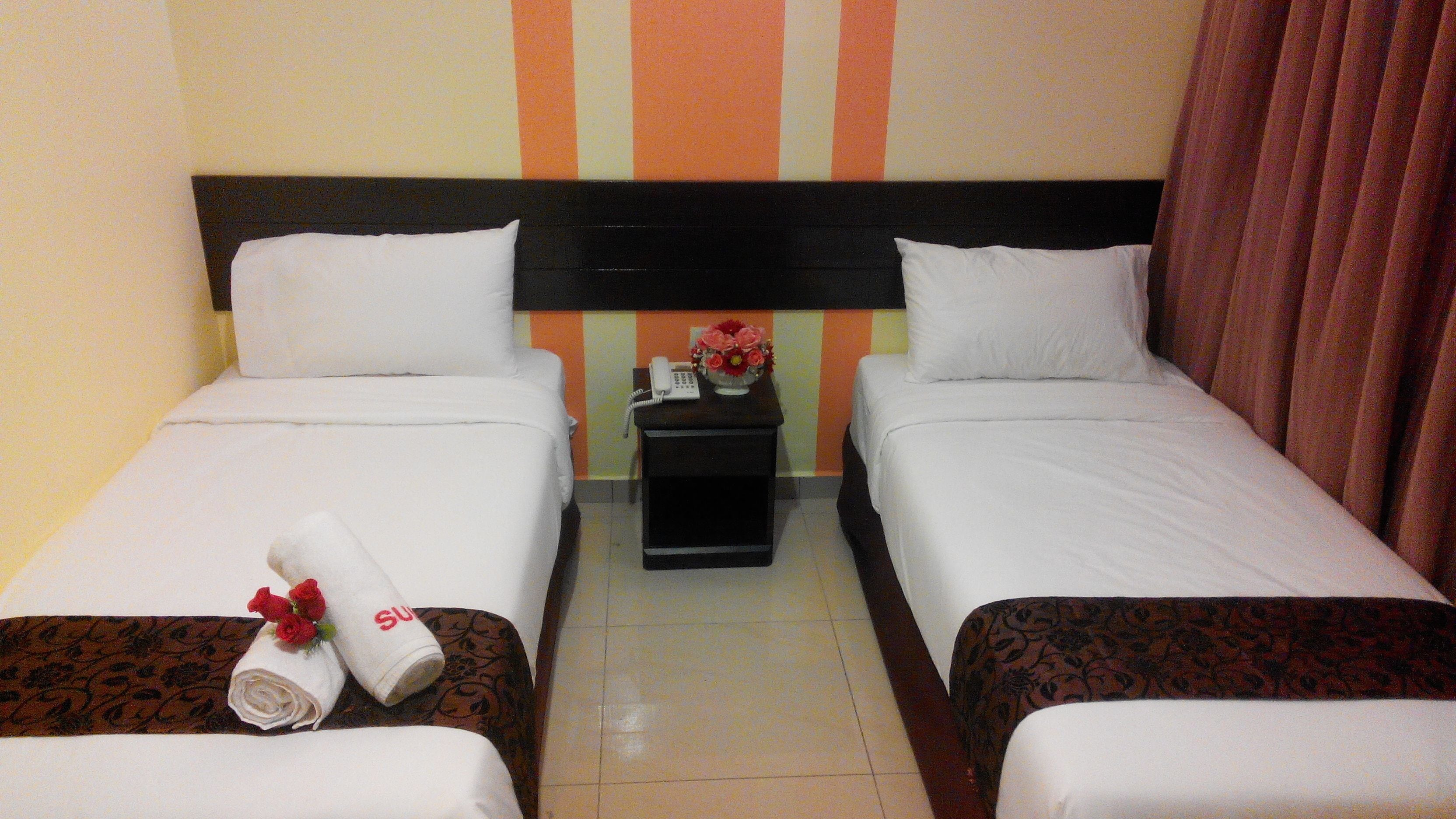 Quarto de hotel com duas camas, uma mesa de cabeceira com telefone e um vaso de flores, e uma parede listrada.