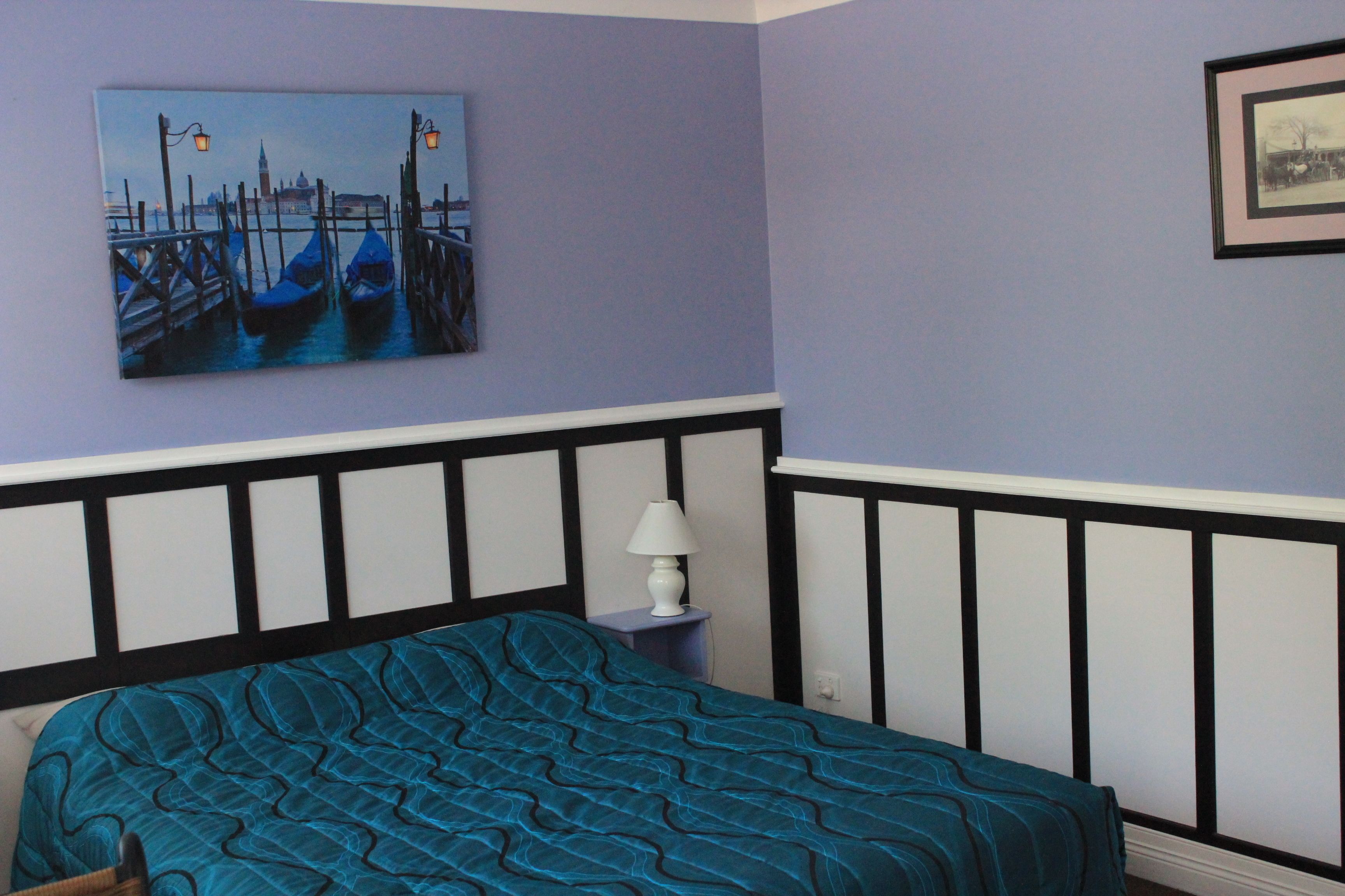 Um quarto com uma cama, uma mesa de cabeceira com abajur, uma pintura de uma cena de canal e uma moldura na parede.
