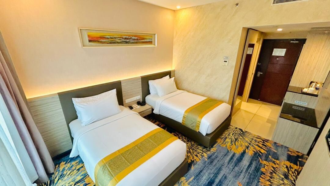 Quarto de hotel com duas camas, uma escrivaninha, uma cadeira, um quadro na parede e uma porta.