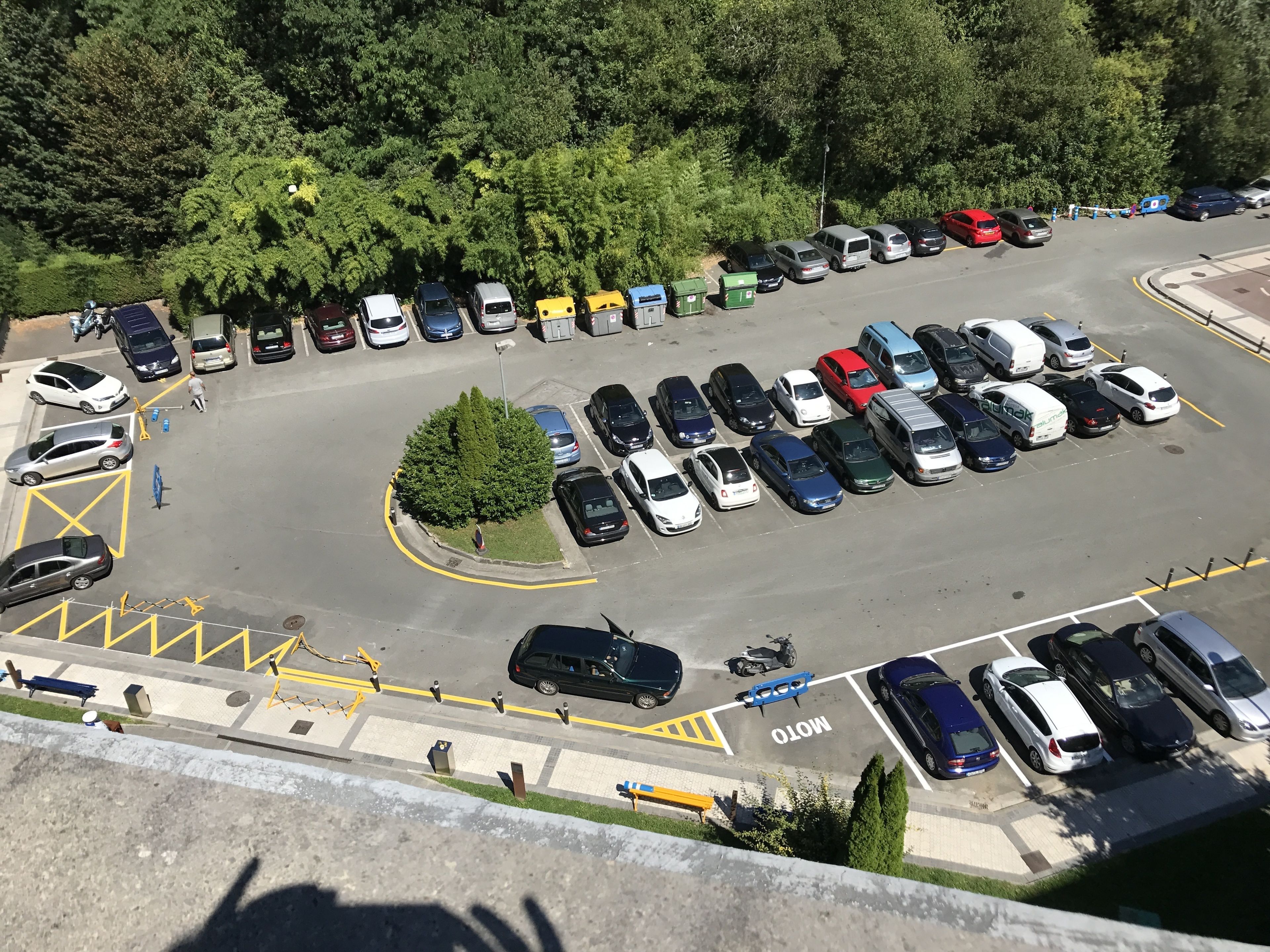 Vista aérea de um estacionamento com várias filas de carros estacionados e uma área verde central.