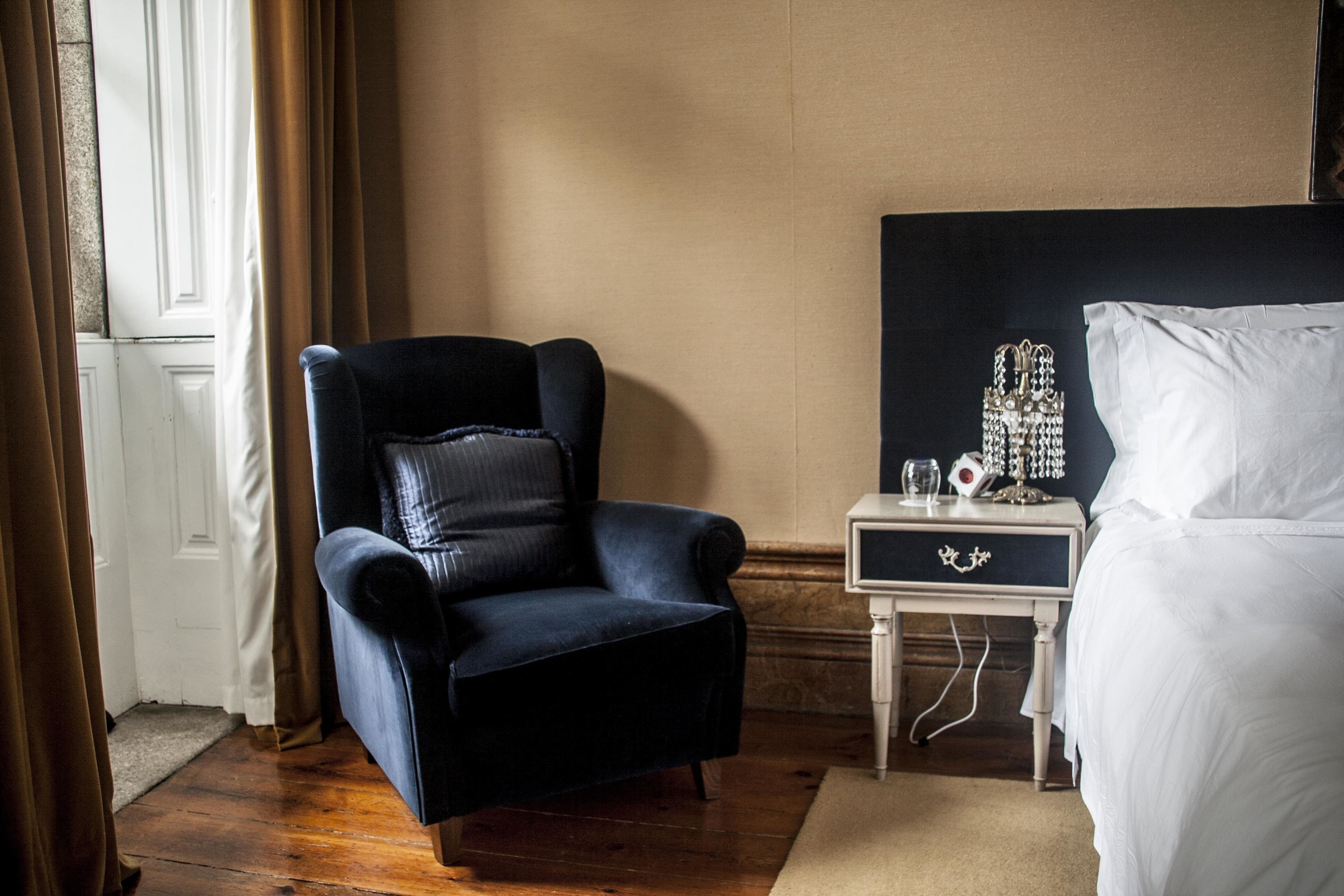 Um quarto com uma poltrona azul escura, uma cama branca, uma mesa de cabeceira com abajur e uma obra de arte na parede.