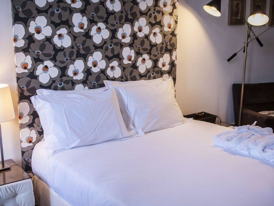 Quarto de hotel com uma cama, almofadas brancas, abajur, mesa de cabeceira, toalha dobrada e cabeceira estampada com flores.
