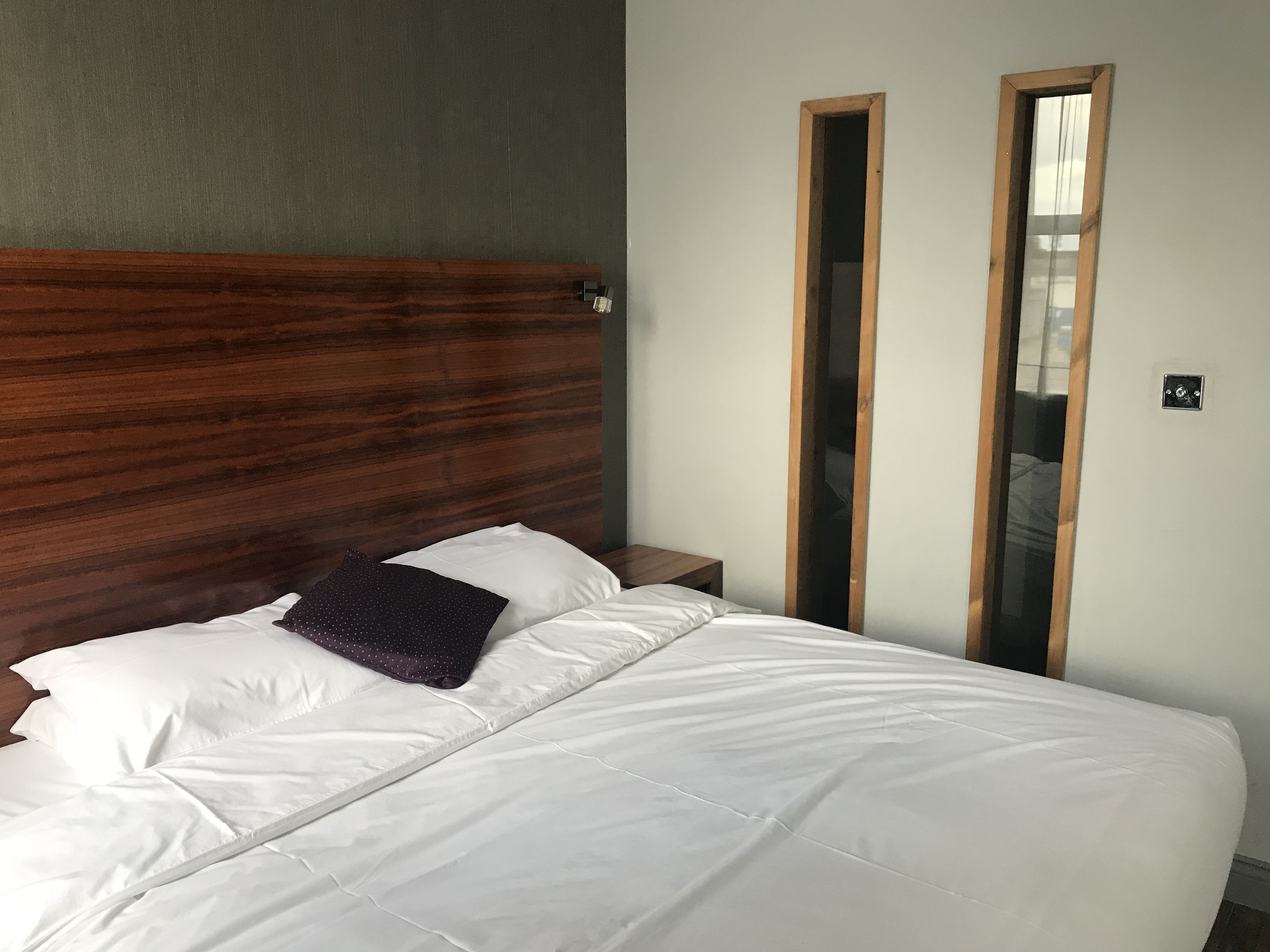Cama bem arrumada com roupa de cama branca e travesseiro escuro, cabeceira de madeira e dois espelhos verticais na parede.