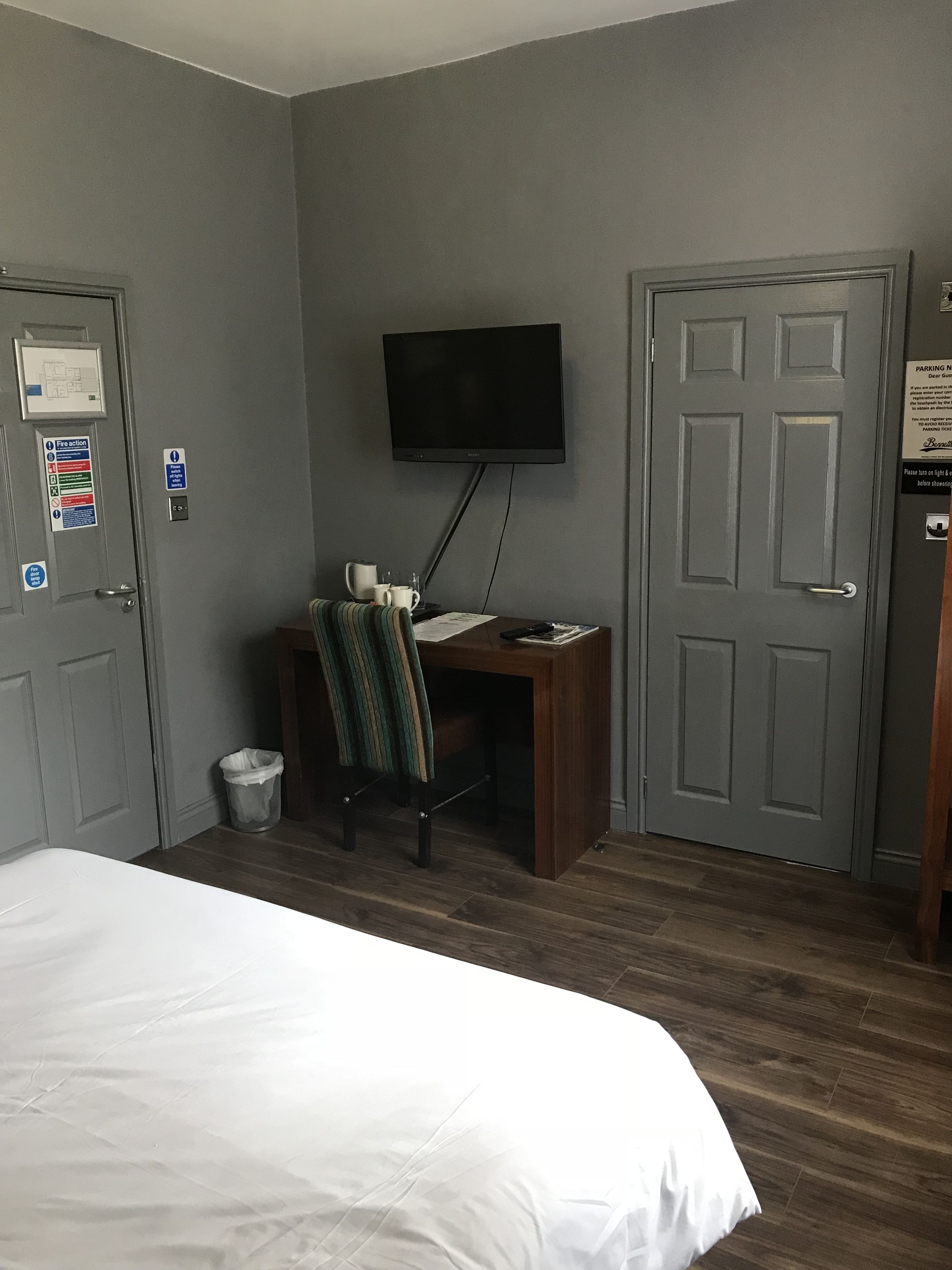 Quarto de hotel com uma cama, uma escrivaninha com cadeira, uma televisão na parede e uma porta com cartaz de procedimentos em caso de incêndio.