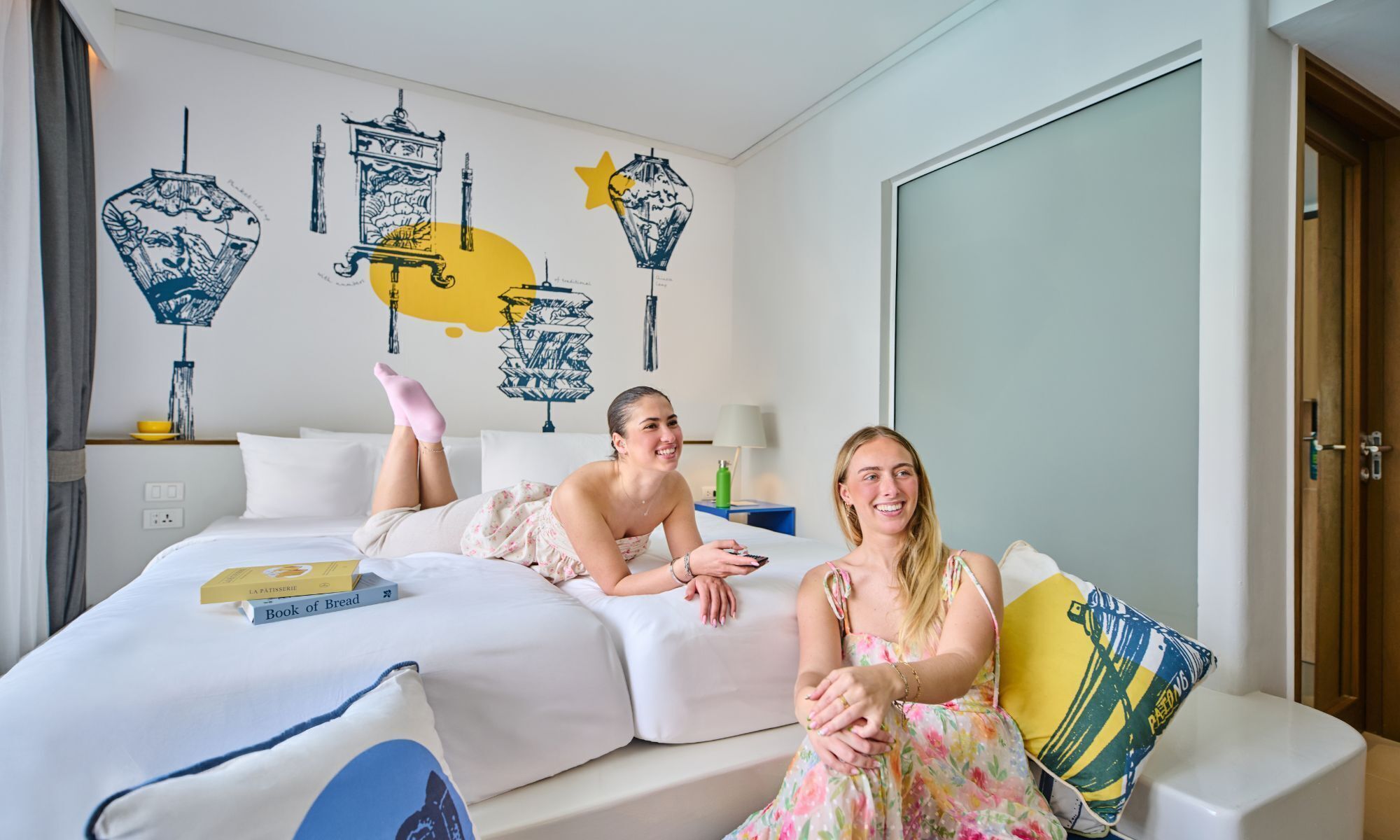 Quarto de hotel com cama, duas pessoas deitadas, mesa de cabeceira com abajur e arte nas paredes.